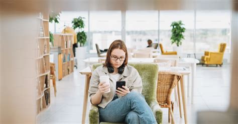 젊은 갈색 머리 십대 소녀 대학 또는 안경을 쓴 고등학생은 녹색 현대 도서관 공공 장소 녹색 열린 공간 사무실에서 일하는 휴대 전화를 사용합니다 사진