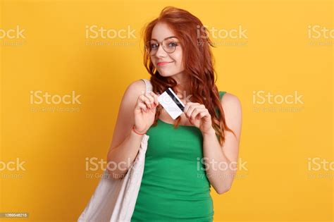 쇼핑백과 신용카드카드를 들고 카메라를 향해 직접 미소짓고 노란 벽에 빨간 머리를 한 여성 쇼핑몰에서 쇼핑을 하고 있는 백인 여성