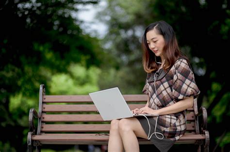 공원 벤치에서 이어폰을 끼고 노트북을 사용하여 나무와 평화로운 녹색 자연으로 둘러싸인 야외에서 원격 근무를 즐기는 미소 짓는 젊은 여성 Central Florida에 대한