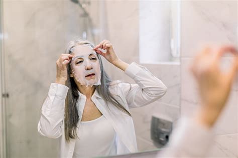 욕실에서 아름다운 회색 머리 은퇴 한 여성의 초상화 그녀의 얼굴에서 흰색 시트 마스크를 제거 집에 주름 보습 뷰티 트리트먼트를 받고 여자 노인에 대한 스톡 사진 및 기타