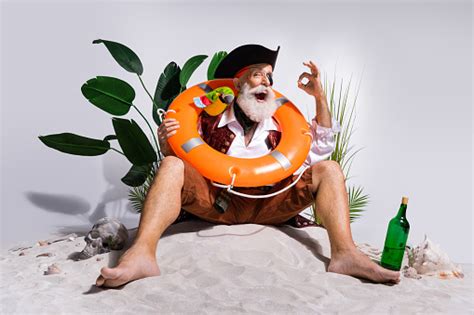 해적 의상을 입은 쾌활한 노인이 구명 부표와 열대 식물과 함께 모래 해변에 앉아 있습니다 55 59세에 대한 스톡 사진 및 기타