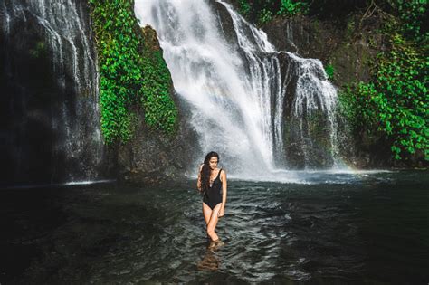 발리에서 거대한 열대 폭포 바뉴말라의 석호에서 즐기는 곱슬 머리와 젊은 슬림 갈색 머리 여자 검은 수영복을 입고 인도네시아에서 행복한 휴가 방랑자 여행 개념입니다 가냘픈에