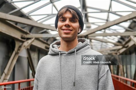 회색 후드티와 흰색 무선 이어버드 헤드폰이 달린 모자를 쓰고 웃는 힙스터 남자의 도시 초상화 남자에 대한 스톡 사진 및 기타 이미지 Istock