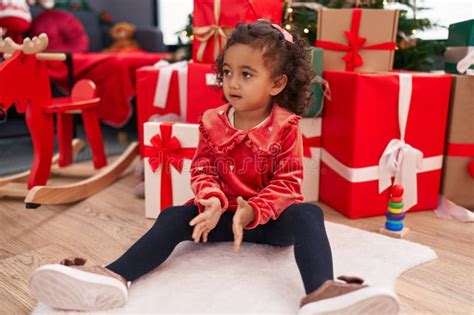 Adorable Hispanic Girl Sitting On Floor By Christmas Tree At Home Stock