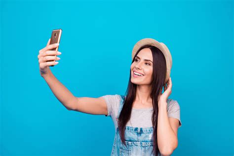 명랑 하 고 젊은 매력적인 갈색 머리는 파란색 바탕에 웃 그녀는 Selfie에 복용 그녀의 휴대 전화의 카메라 캐주얼 여름 옷과