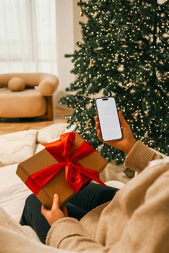 아늑한 휴가 환경에 앉아 빈 화면이 있는 스마트폰과 빨간 리본으로 아름답게 포장된 선물 상자를 들고 있는 사람 Brand Name Smart Phone에 대한 스톡 사진 및