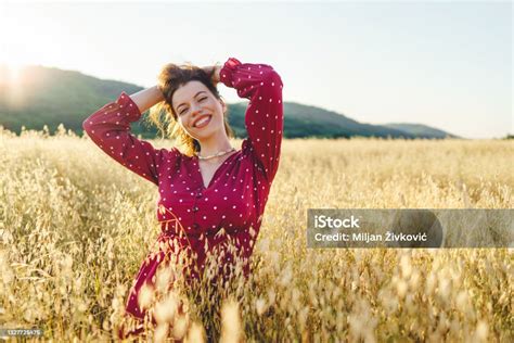 여름날에 빨간 드레스를 입은 성인 백인 갈색 머리 여성의 초상화는 복사 공간으로 카메라를 보고 행복한 미소를 짓는 작물 곡물 밭에 서 있습니다 20 24세에 대한 스톡 사진