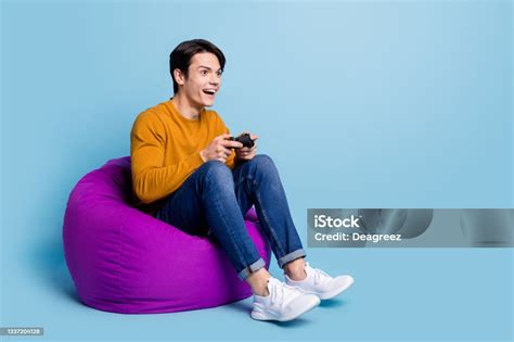 블루 컬러 배경에 고립 된 가방 플레이 스테이션 착용 스웨터 청바지에 앉아 감동 브루넷 젊은 남자의 전체 길이 사진 게이머에 대한 스톡 사진 및 기타 이미지 Istock