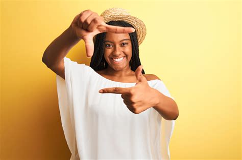 흰색 티셔츠와 모자를 쓰고 고립된 노란색 배경 위에 행복한 얼굴로 손과 손가락으로 프레임을 만드는 젊은 아프리카 계 미국인 여성 창의력과 사진 개념 땋은 머리에 대한 스톡