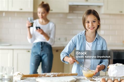 엄마의 작은 도우미 귀여운 백인 어린 소녀는 파이를 요리하는 법을 배운다 계란을 친다 카메라를보고 미소 엄마는 전화를 사용하여 배경에 서 2명에 대한 스톡 사진 및 기타