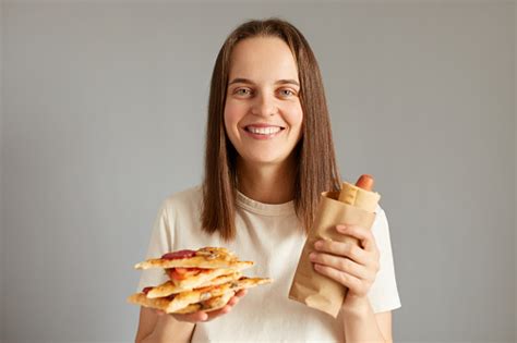 회색 배경에 격리된 핫도그와 피자 조각을 들고 패스트푸드를 즐기며 즐거운 쾌활한 표정으로 카메라를 바라보는 어린 예쁜 소녀의 초상화 30 34세에 대한 스톡 사진 및 기타