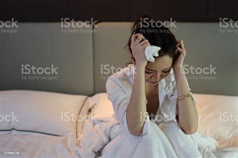 캐주얼 드레스를 입은 젊은 우울한 아시아 여성은 낮은 조명 환경의 어두운 침실에서 침대에서 울면서 손으로 머리를 잡고 앉아 있다