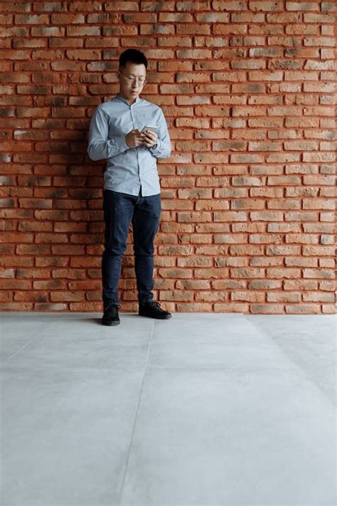 A Man In Blue Long Sleeves And Denim Pants Standing Near Brick Wall