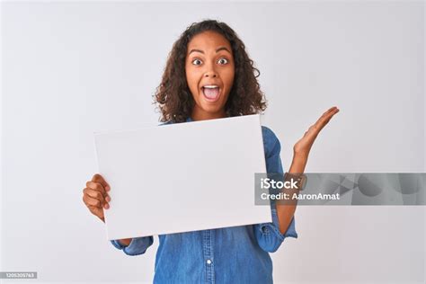 고립된 흰색 배경 위에 현수막을 들고 있는 젊은 브라질 여성은 매우 행복하고 흥분하며 큰 미소와 손을 들고 비명을 지르는 승리를