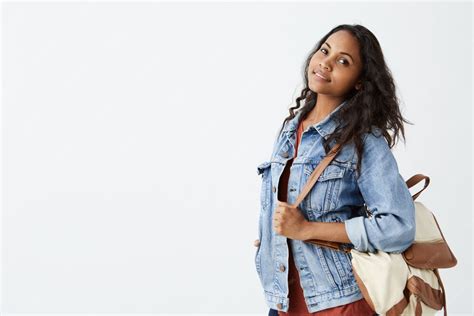 벽에 포즈를 취하는 그녀의 어깨에 배낭과 데님 재킷과 빨간 티셔츠에 검은 물결 모양의 머리 사랑스러운 미소와 아름 다운 긍정적 인 친절한 찾고 젊은 아프리카 계 미국인 여자