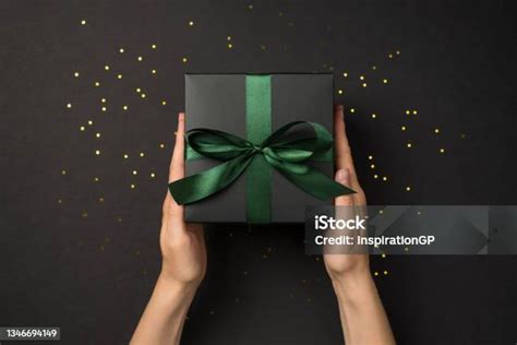 고립된 검은 색 배경에 녹색 리본 활과 황금 색종이가 달린 검은 선물 상자를 들고 있는 여성 손의 1인칭 탑 뷰 사진 녹색에