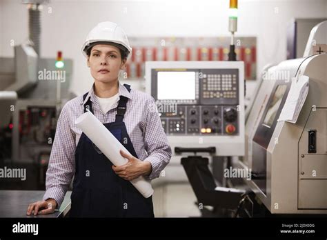 Portrait Of Serious Confident Female Engineer In Hardhat Standing At Cnc Machine And Holding
