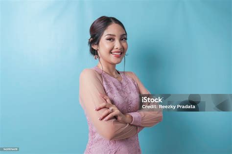 팔을 접은 채 서서 파란 배경 위에 고립된 카메라를 바라보며 모던한 케바야 드레스를 입고 서 있는 자신감 넘치는 웃는 소녀의