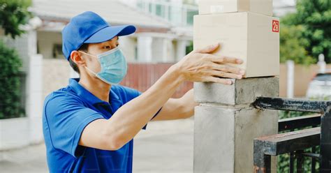 젊은 우편 배달 택배 남자는 집에서 고객에게 보내기 위해 소포 상자를 취급하는 안면 마스크를 착용하고 아시아 여성은 야외에서