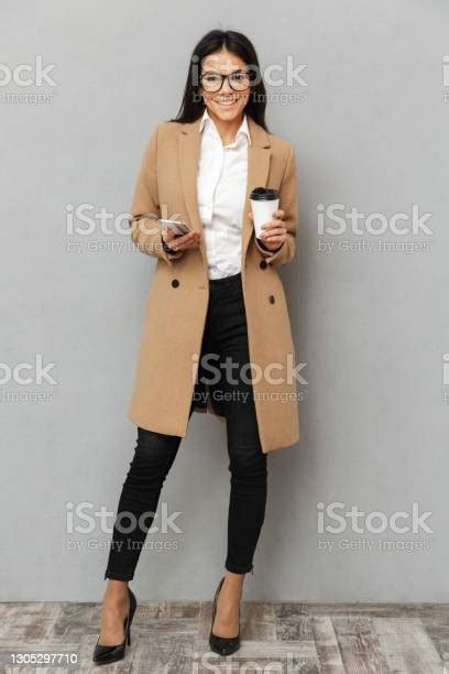 회색 배경에 고립 된 휴대 전화와 테이크 아웃 커피를 손에 들고 서있는 아우터웨어를 입은 아름다운 여성의 전체 길이 이미지