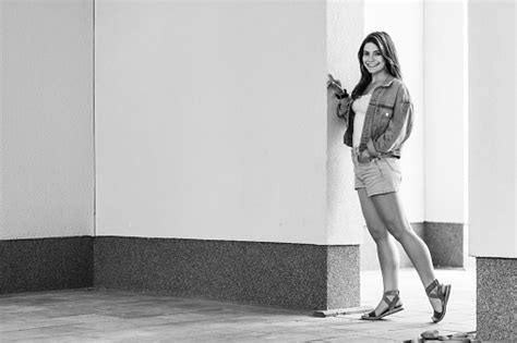 아름 다운 행복 한 갈색 머리 젊은 여자의 흑백 전체 길이 초상화 서 서 벽에 기대어 포즈와 당신의 미소와 함께 카메라를 보고 갈색 머리에 대한 스톡 사진 및 기타 이미지