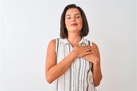 편안한 흰색 배경 위에 서있는 캐주얼 스트라이프 셔츠를 입은 젊은 아름다운 여성은 닫힌 눈과 얼굴에 감사하는 제스처로 가슴에 손을 대고 미소 짓고 있습니다 건강 개념 갈색