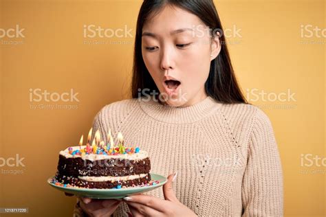 노란 고립 된 배경에 불타는 촛불과 함께 생일 케이크를 들고 있는 젊은 아시아 여성은 깜짝 얼굴로 충격에 무서워하고 두려움과 두려움표현으로 흥분했습니다 생일 케이크에 대한