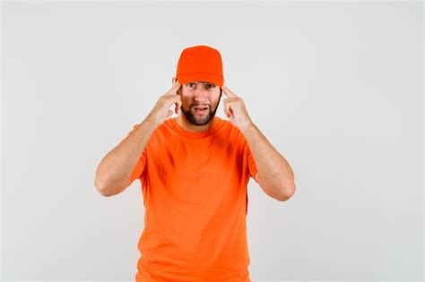 주황색 티셔츠를 입은 배달원 관자놀이에 손가락을 대고 현명하게 보이는 모자 앞모습 무료 사진