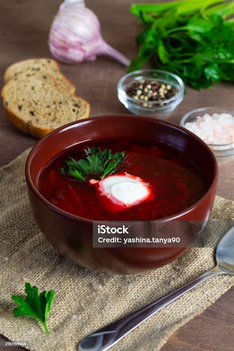 붉은 사탕무 수프 숟가락이 있는 갈색 수프 그릇 사워 크림 마늘 그린 파슬리를 곁들인 맛있는 보르시 나무 배경에 전통적인 러시아와 우크라이나 요리 복사 공간 세로 갈색에
