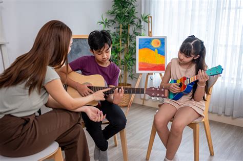 초등학교 기타 교실에서 학생들을 교육하는 아시아계 교사 열정적인 여성 교사가 교실에서 의자에 앉아 기타를 열심히 연주하는 귀여운 아이들에게 수업을 설명합니다 10 11세에