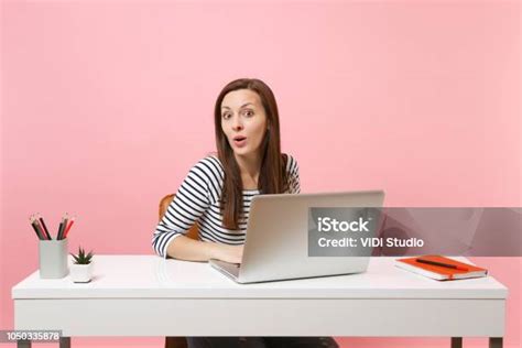 영 캐주얼 옷 프로젝트 작업을 노트북으로 사무실에 앉아 파스텔 핑크 배경에 고립 동안에 여자를 충격 성과 비즈니스 경력 개념입니다 광고에 대 한 공간을 복사 합니다 갈색