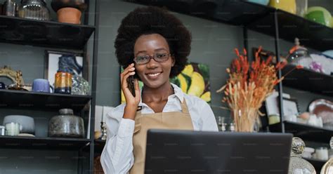 예쁜 아프리카 판매원이 모바일로 이야기하고 장식 가게에서 노트북으로 입력하는 모습을 닫습니다 선반에 누워있는 다양한 아름다운 장식 작업을위한 현대 가제트 사진