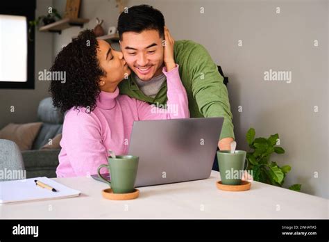 Multiethnic Happy Couple Kissing On A Cheek While Using The Laptop