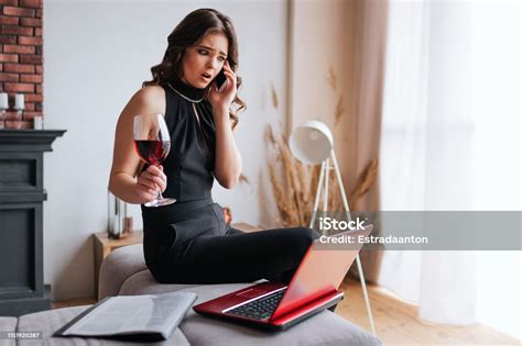 젊은 사업가 집에서 작동 합니다 테이블에 앉아서 전화로 얘기 손에 레드 와인 잔을 들고 갈색 머리 모델 착용 블랙 드레스와 갈색