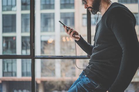 젊은 사람이 서 고 듣는 디지털 오디오도 서에 대 한 휴대 전화를 사용 하 여 수염 Horizontalpanoramic 창 흐린