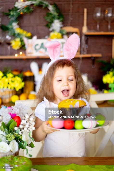 부활절 부활절 케이크와 화려한 페인트 달걀을 가진 어린 소녀가 그것을 먹고 부엌에서 집에있는 축제 테이블에서 집에서 토끼의 귀를 핥습니다 2 3 살에 대한 스톡 사진 및