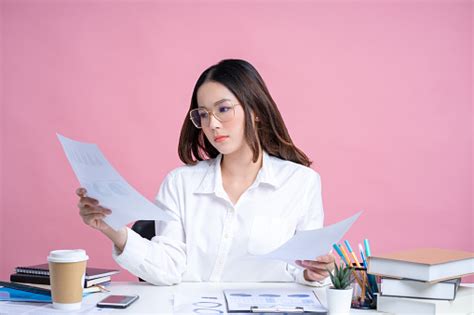 안경을 쓰고 흰 셔츠를 입은 젊은 아시아 여성이 흰색 책상에 앉아 있다 그녀는 사무실에서 노트북으로 문서를 들고 있다 N 파스텔