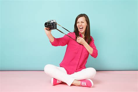 전체 길이 초상화 밝은 분홍색 파란 파스텔 벽 배경 스튜디오에 고립 된 바닥 홀드 카메라에 앉아 장미 셔츠 블라우스 흰색 바지에 젊은 여자 패션 라이프 스타일 개념입니다