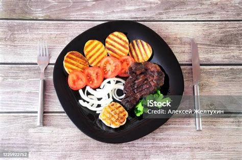 감자 토마토 양파를 곁들인 소고기 스테이크는 검은 색 유리 접시와 칼붙이위에 나무 테이블에 올려 놓습니다 0명에 대한 스톡 사진