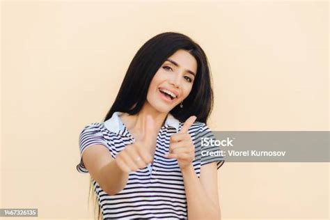 매력적인 유럽 여성의 샷은 두 엄지 손가락을 주고 긍정적 인 표정을 가지고 있으며 브래킷이있는 흰색 치아를 가지고 있으며 캐주얼하게 옷을 입고 괜찮아지고 승인 제스처를