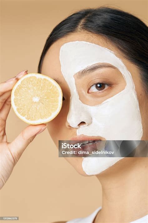 얼굴 마스크를 쓴 젊은 여성의 뷰티 초상화가 레몬 조각을 들고 베이지 색 배경위에 고립된 포즈를 취하는 얼굴의 절반에 가세합니다 18 19세에 대한 스톡 사진 및 기타