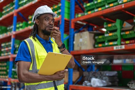 조끼와 헬멧을 착용한 흑인 남성 창고 노동자가 휴대용 무전기를 이용해 창고 공장 매장에서 물건을 확인하면서 동료와 통화하고 있다 물류 물류센터 개념 30 34세에 대한 스톡