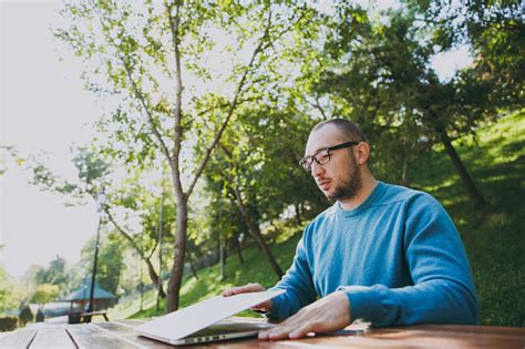 영 성공적인 스마트 남자 사업 또는 캐주얼 블루 셔츠 안경 녹색 자연에 야외에서 일 하는 노트북을 사용 하 여 도시 공원에서 휴대 전화와 함께 테이블에 앉아 있는 학생