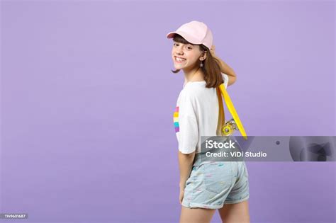 생생한 옷에 쾌활 한 십 대 소녀의 초상화 스튜디오에서 보라색 파스텔 배경에 고립 된 노란 스케이트 보드를 들고 있습니다
