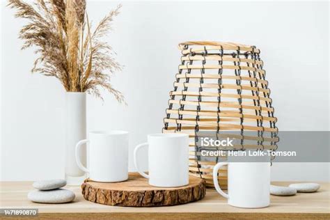 세 개의 흰색 빈 머그잔이 조롱합니다 갈대와 장식용 나무 랜턴이 있는 흰색 꽃병이 있는 나무 테이블에 여러 컵이 있습니다 빈