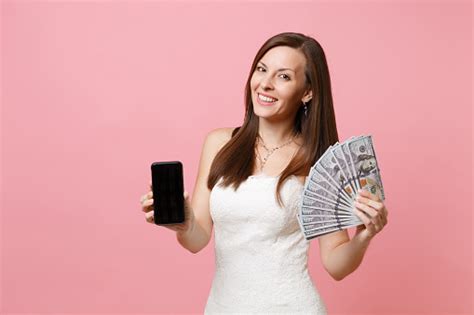 웨딩 드레스 번들 많은 달러 현금 돈 분홍색 배경에 고립 빈 검은색 빈 화면으로 휴대 전화를 들고 웃는 신부 여자 결혼식 축