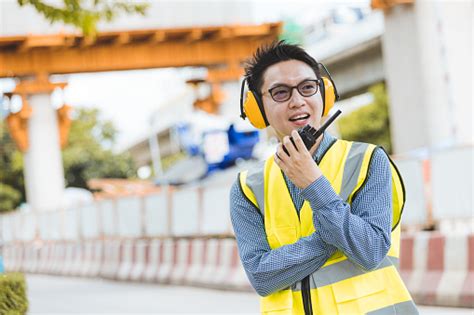노란색 보호 귀 머프의 산업 건설 엔지니어 외부 이미지는 크레인 근처의 열린 건물 현장에서 무전기와 행복한 미소를 사용하면서