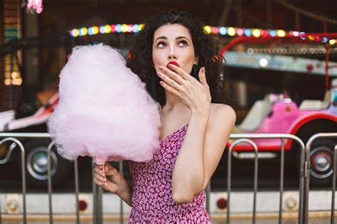 분홍색 면사탕을 손에 들고 서있는 드레스에 어두운 곱슬 머리를 가진 아름다운 소녀는 놀이 공원에서 시간을 보내는 동안 꿈에서 옆으로 쳐다 보고 솜사탕에 대한 스톡 사진 및