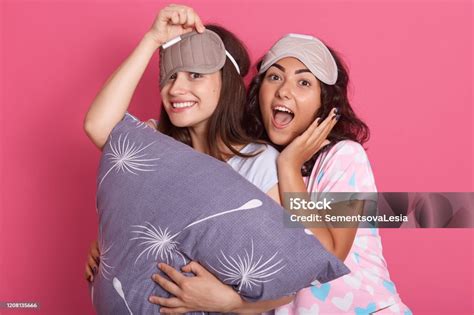 기쁘게 보이는 매력적인 젊은 갈색 머리의 그림 베개를 들고 잠옷을 입고 머리에 마스크를 자고 스튜디오에서 분홍색 배경 위에 고립 된 서있습니다 감정에 대한 스톡 사진 및