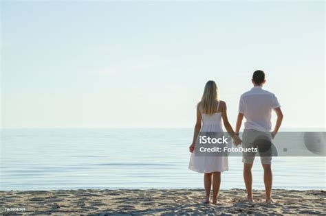 하얀 옷을 입은 젊은 부부는 모래 위에 서서 잔잔한 바다와 밝은 푸른 하늘을 바라보고 있습니다 신선한 공기 감상적인 영감을 주는